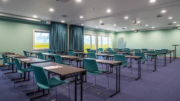 Uma sala de conferências com fileiras de mesas e cadeiras, janelas amplas com cortinas e uma tela de projeção.