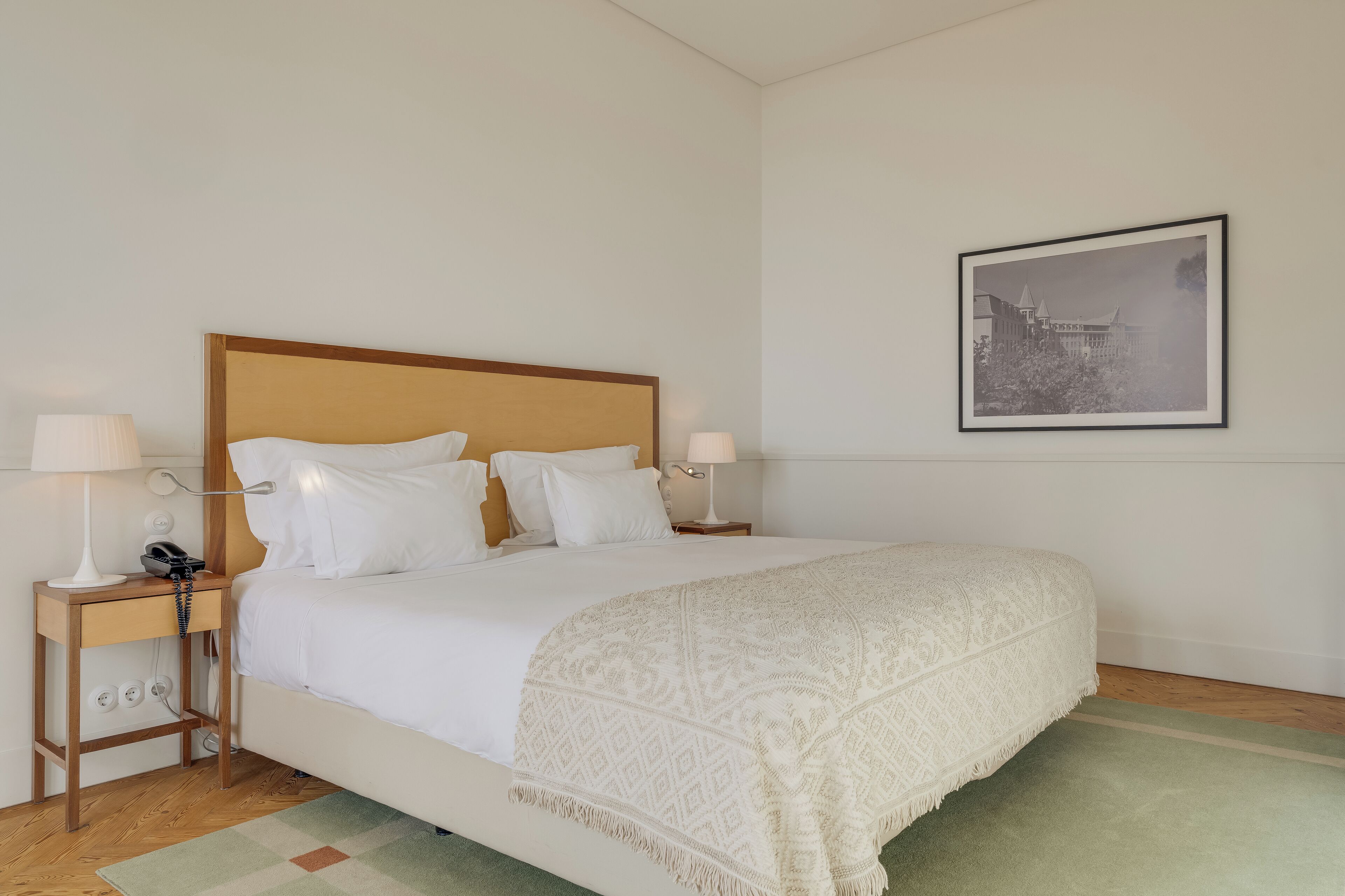 Uma cama bem arrumada com cabeceira de madeira, duas mesas de cabeceira com abajures, um cobertor estampado e um quadro na parede.