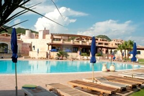 Piscina com espreguiçadeiras e guarda-sóis, cercada por edifícios com arquitetura mediterrânea.