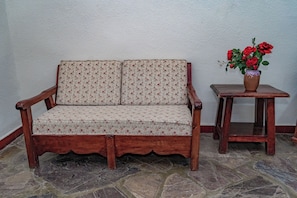 Um banco de madeira com almofada estampada, uma pequena mesa de madeira com um vaso de flores vermelhas e um chão de azulejos de pedra.