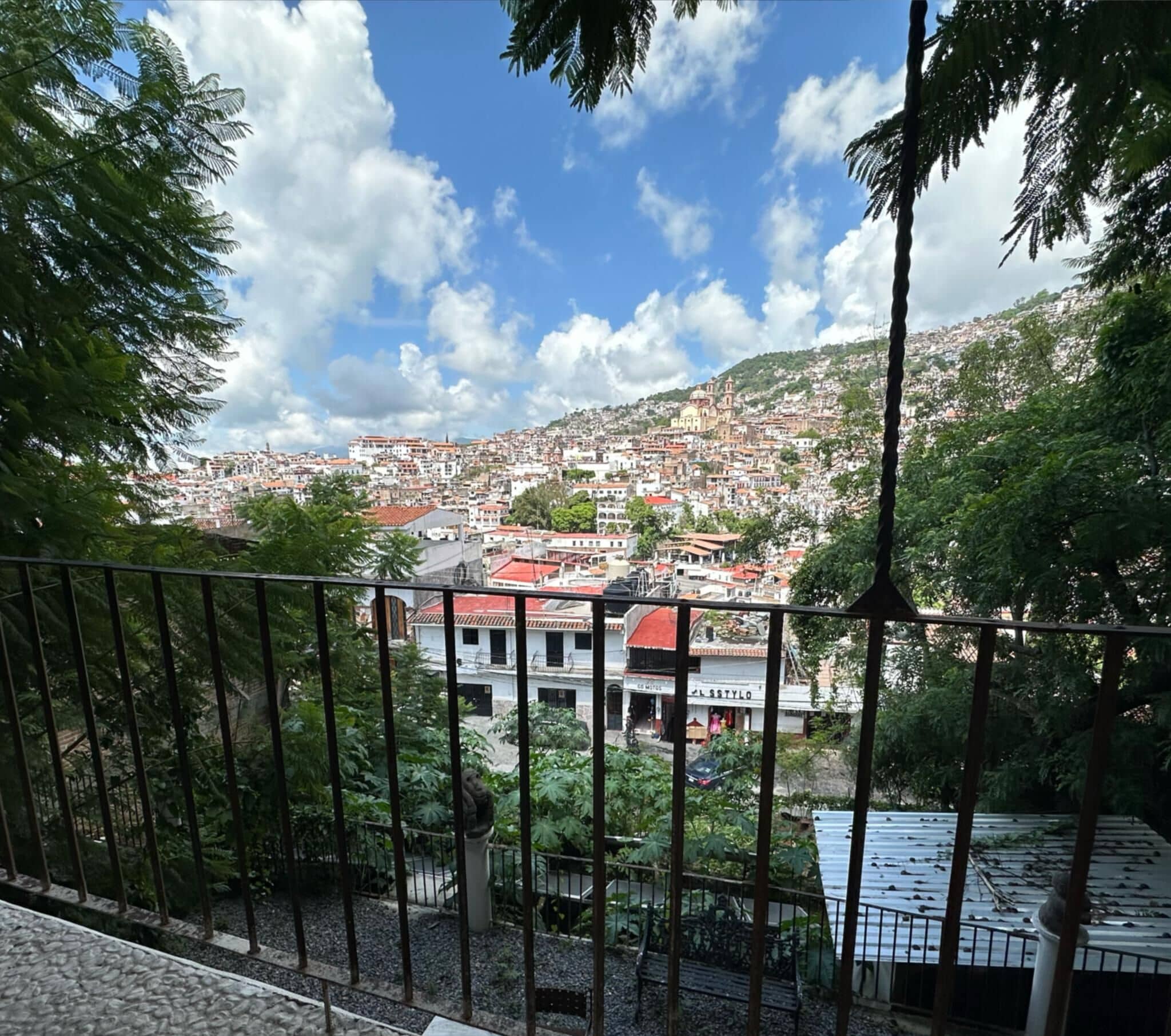 Uma varanda com vista para uma cidade em encosta, com uma mistura de edifícios residenciais e comerciais, e uma igreja com uma torre de sino em destaque.