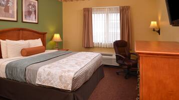 Quarto de hotel com cama, escrivaninha, cadeira, abajur e televisão.