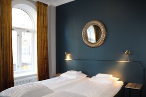 Quarto de hotel com cama grande, espelho redondo e janela com cortinas.