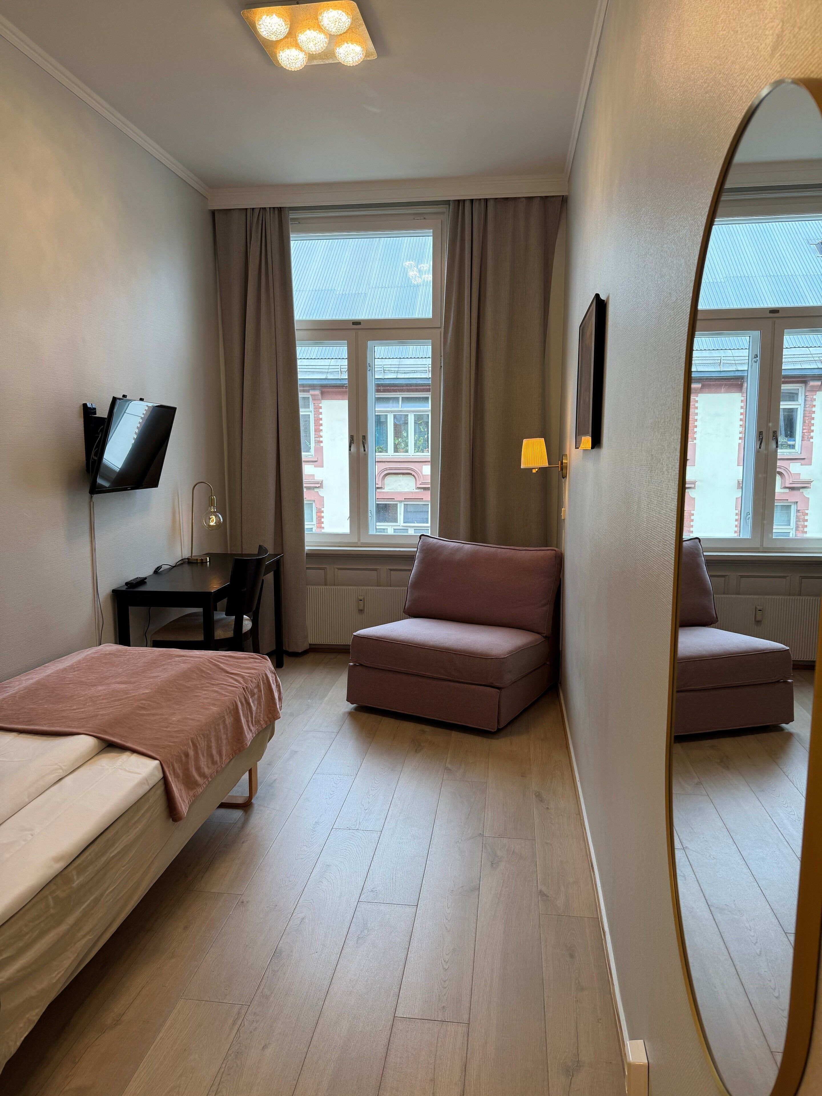 Quarto de hotel com cama, escrivaninha, poltrona, sofá, televisão fixada na parede e um espelho grande.