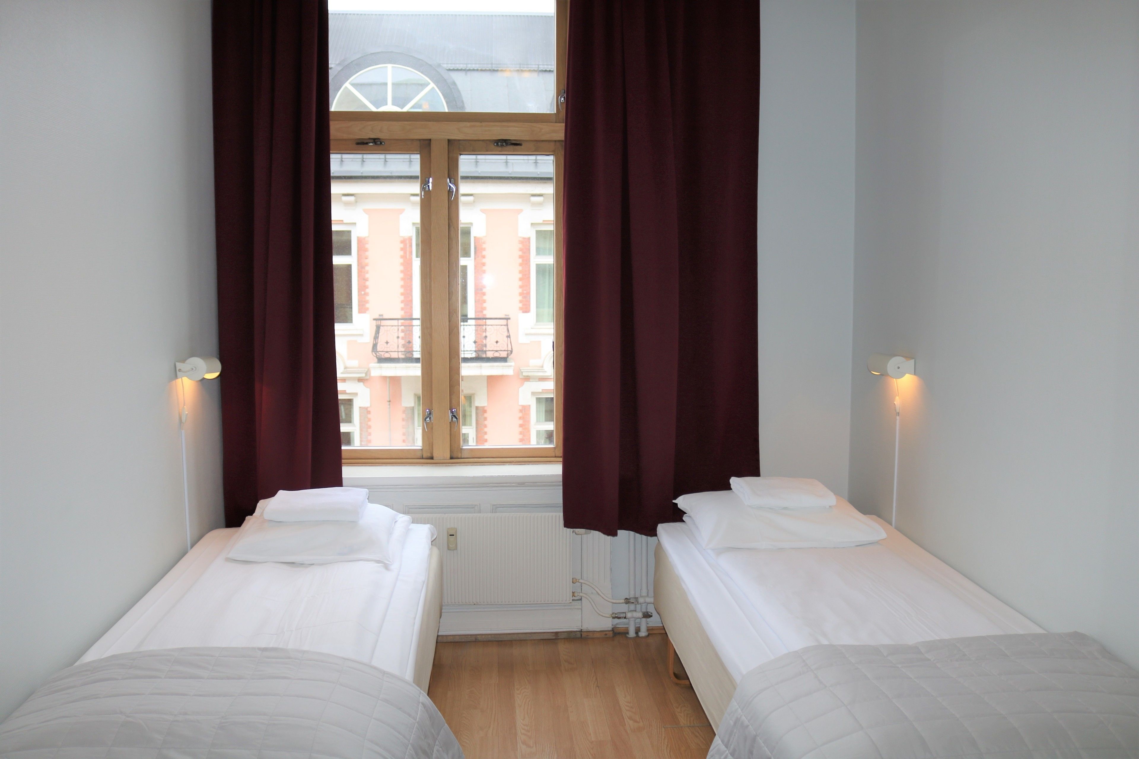 Quarto de hotel com duas camas de solteiro, janela com cortinas, radiador e vista para um edifício.