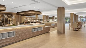 Um lobby moderno de hotel com área de buffet central, grandes pilares de madeira e um espaço de estar com mesas e cadeiras.