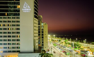 Um hotel com uma placa visível, estacionamento e uma rua com veículos e postes de iluminação.