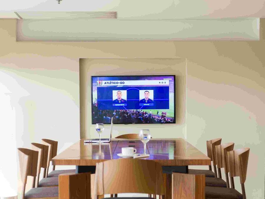 Uma sala de conferência com uma mesa comprida e seis cadeiras, uma TV de tela plana na parede exibindo um evento esportivo, e um teto com iluminação embutida.