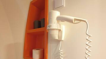 Um telefone fixado na parede com fio enrolado, uma prateleira pequena com um rolo de papel higiênico e uma placa de interruptor branca.