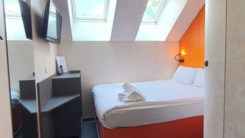 Quarto de hotel pequeno com cama, escrivaninha, televisão e claraboia.