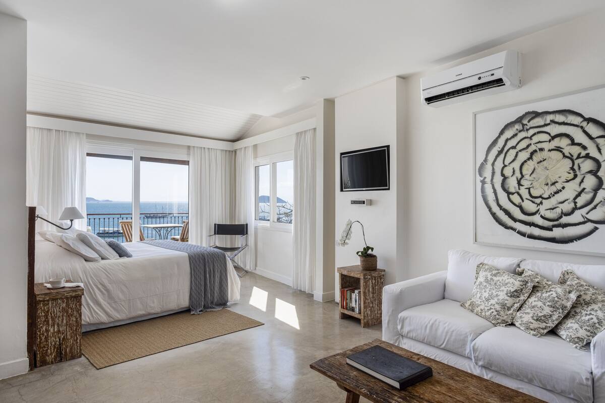 Quarto de hotel moderno com cama, sofá, televisão e uma ampla janela com vista para o mar.