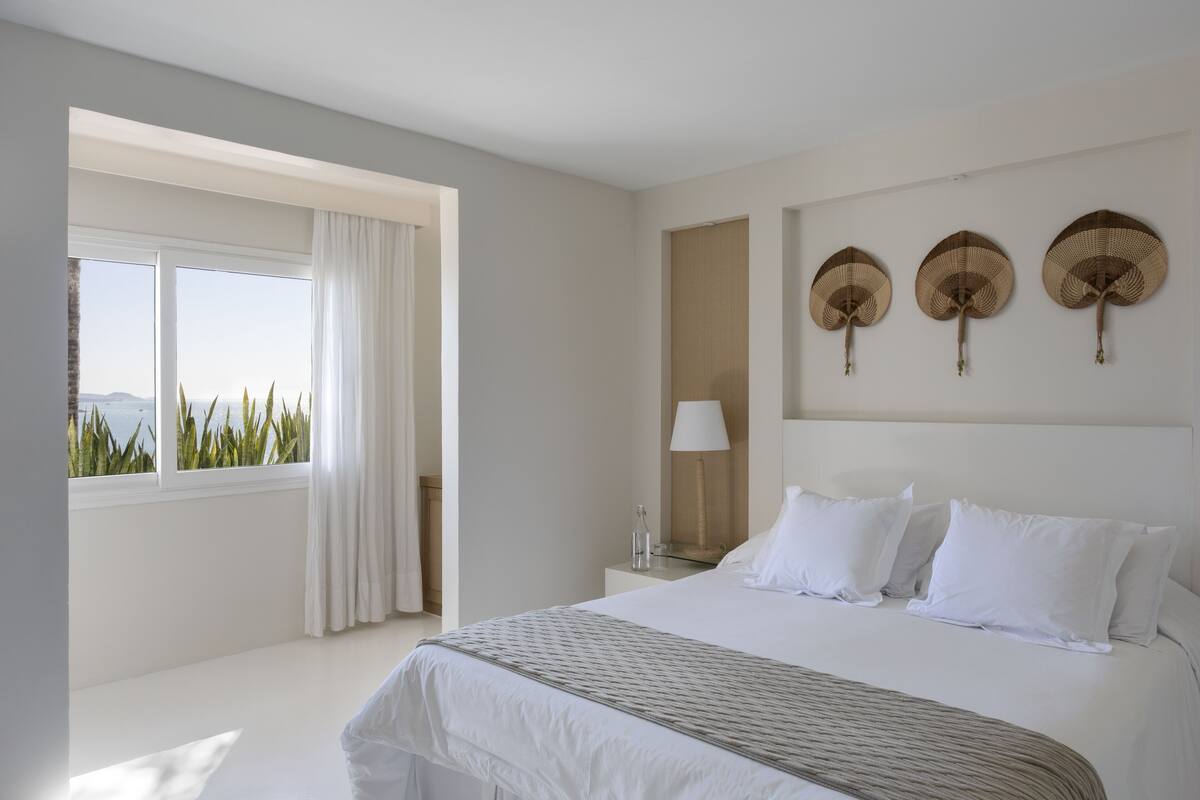 Quarto de hotel moderno com uma cama grande, mesa de cabeceira com abajur e vista para o mar por uma janela ampla.