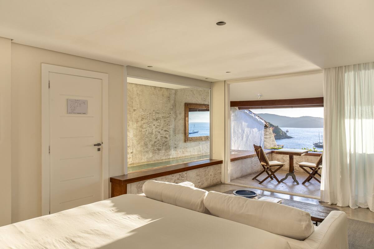 Quarto de hotel com cama, área de estar com mesa e cadeiras, vista para o mar e porta com placa.