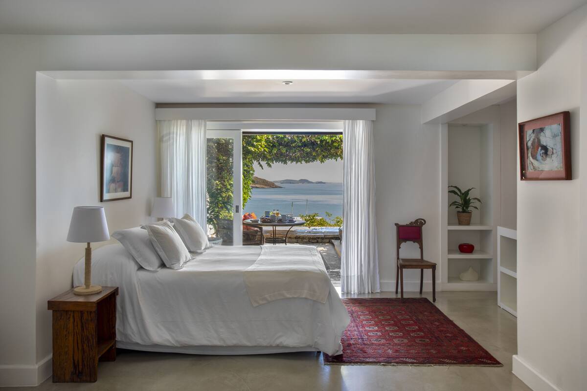 Quarto com cama, mesa de cabeceira, cadeira, mesa e vista para o mar.