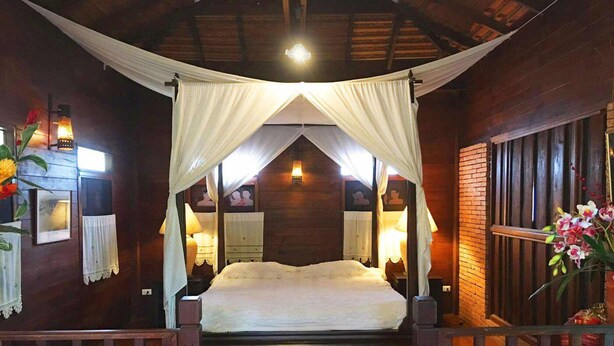 Quarto de madeira com cama sob um dossel, paredes de madeira e uma pequena mesa com abajur.