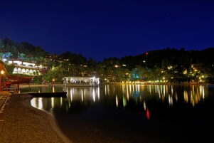 Vista noturna à beira do lago, com um barco atracado na margem, uma ponte e edifícios iluminados ao fundo.