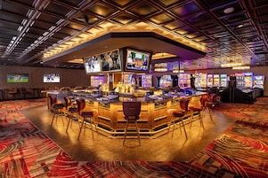 Uma área de bar com várias máquinas de jogo, uma tela grande exibindo esportes e um balcão com banquetas.