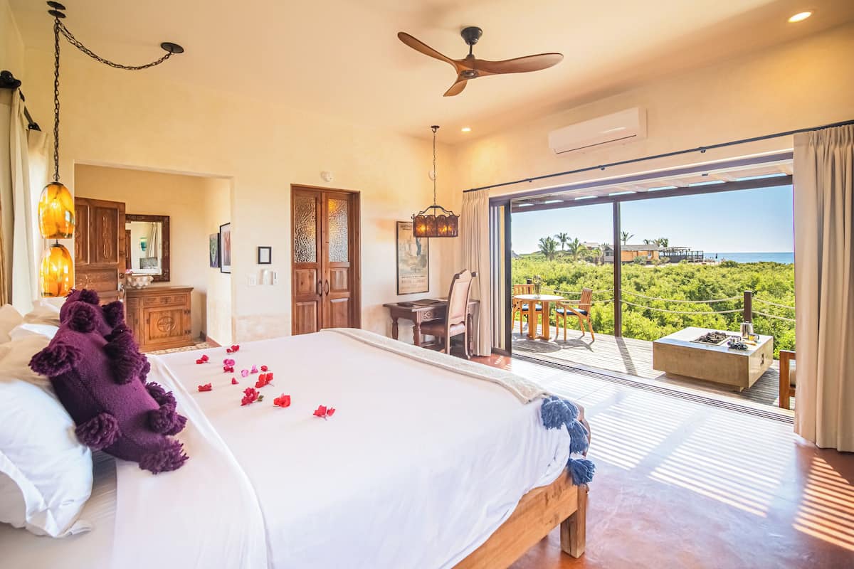 Quarto com cama, cômoda de madeira, escrivaninha e cadeiras, e varanda com vista para a paisagem.