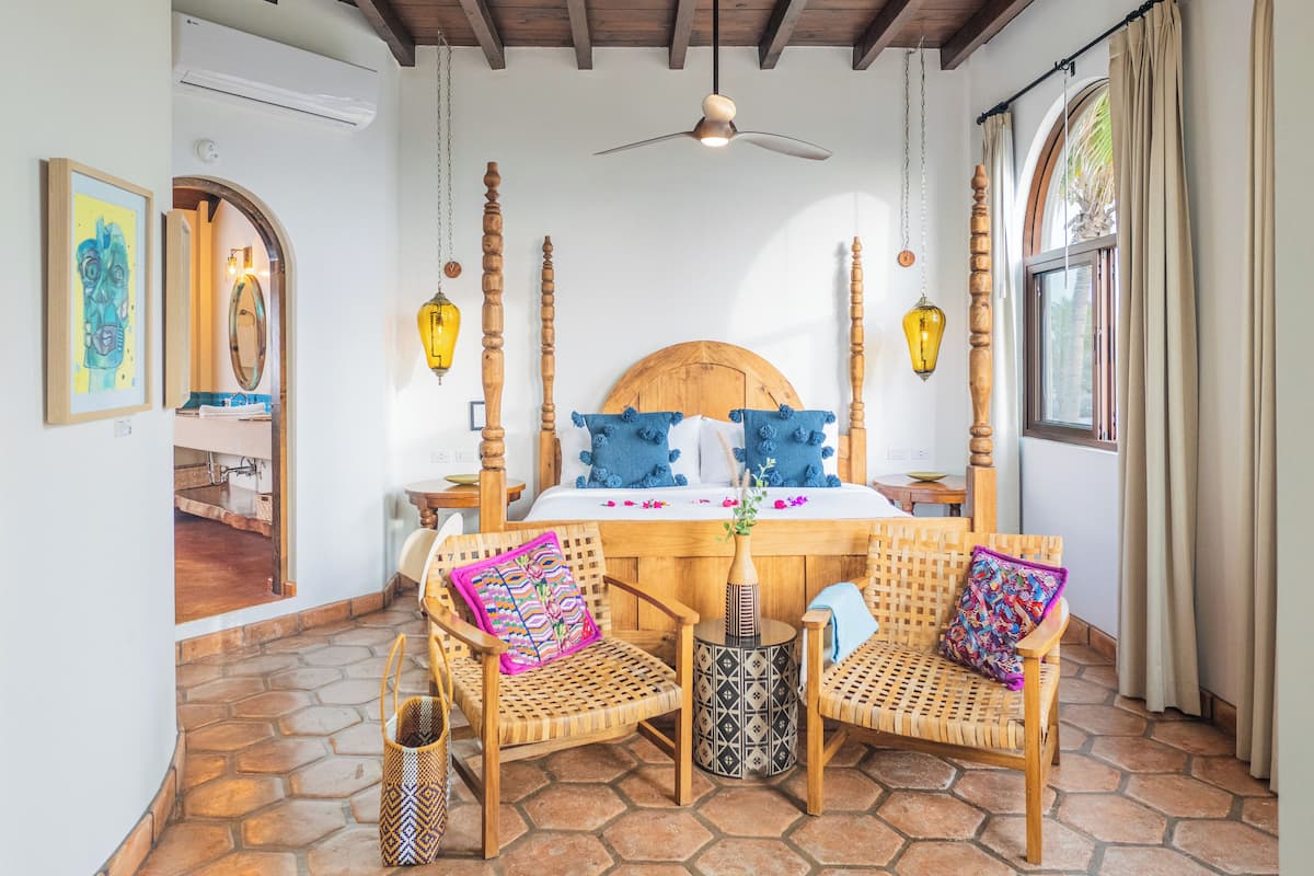 Quarto com teto de madeira, cama com almofadas azuis, cabeceira de madeira, mesa redonda com cadeiras e janela com cortinas.