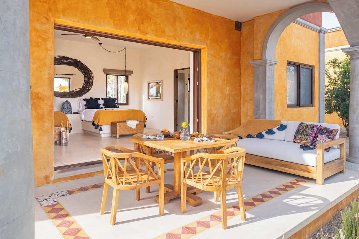Uma sala de estar aconchegante com uma mesa de madeira e cadeiras, um sofá com almofadas estampadas e vista para o quarto adjacente.