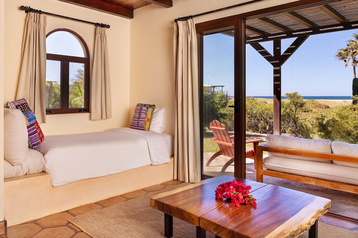 Quarto com cama, sofá, mesa de madeira com arranjo floral e vista para a praia através de grandes janelas.