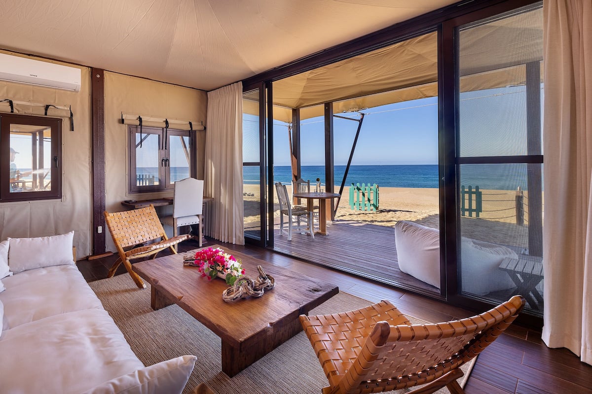 Quarto com uma grande porta de vidro deslizante que dá para a praia, um sofá, uma mesa de madeira com um vaso de flores e cadeiras de vime.
