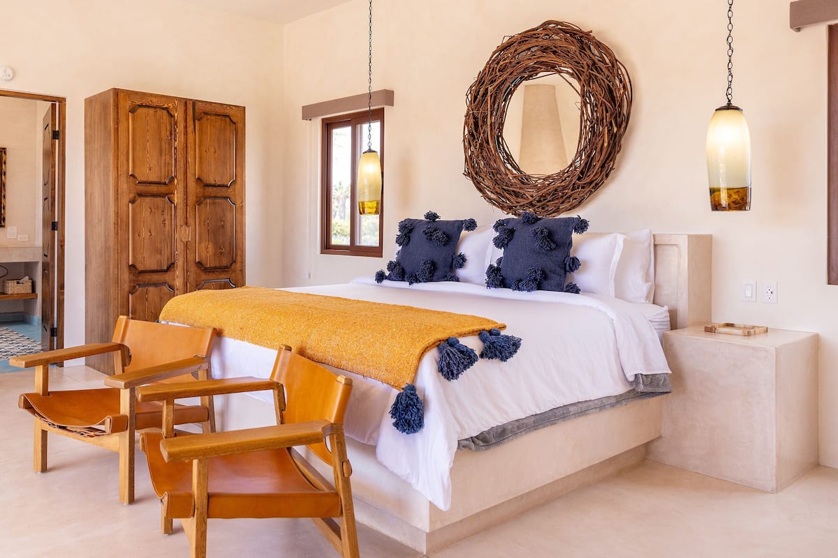 Quarto com cama, duas cadeiras, guarda-roupa de madeira, espelho e luminária pendente.