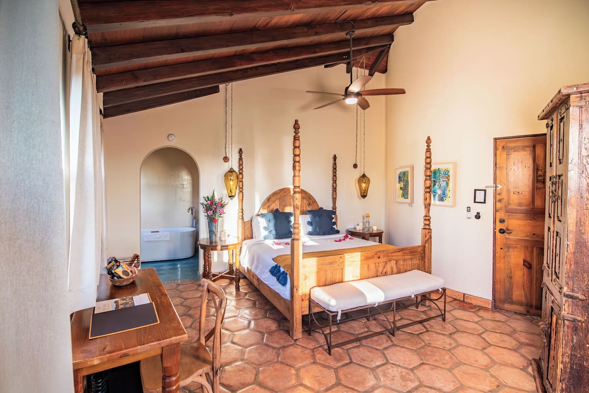 Quarto com cama de dossel, escrivaninha de madeira, um banco, luminária pendente e porta com moldura de madeira.