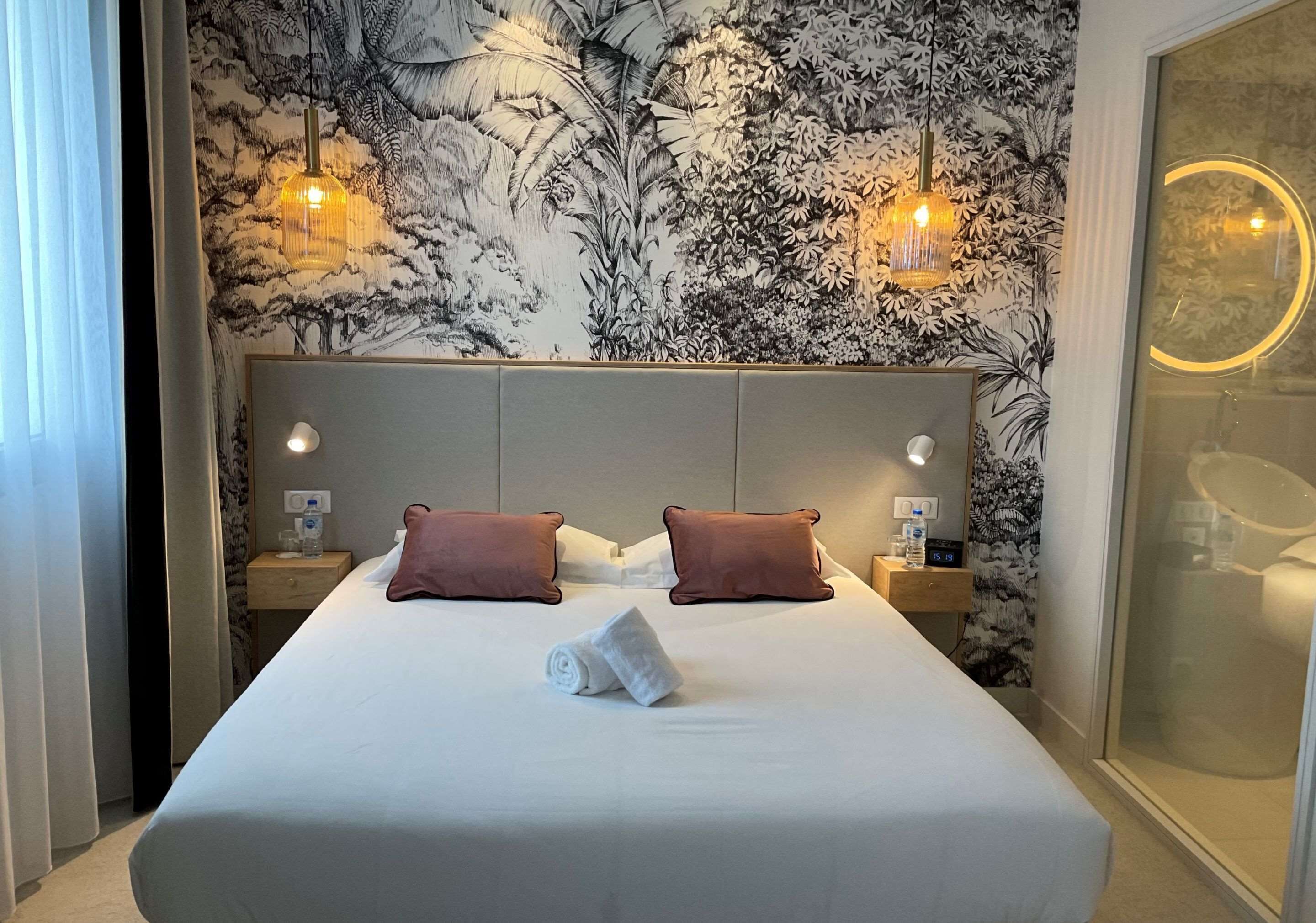 Quarto de hotel com uma cama grande, duas almofadas, uma toalha enrolada, mesas de cabeceira com abajures e uma parede com padrão de folhas.
