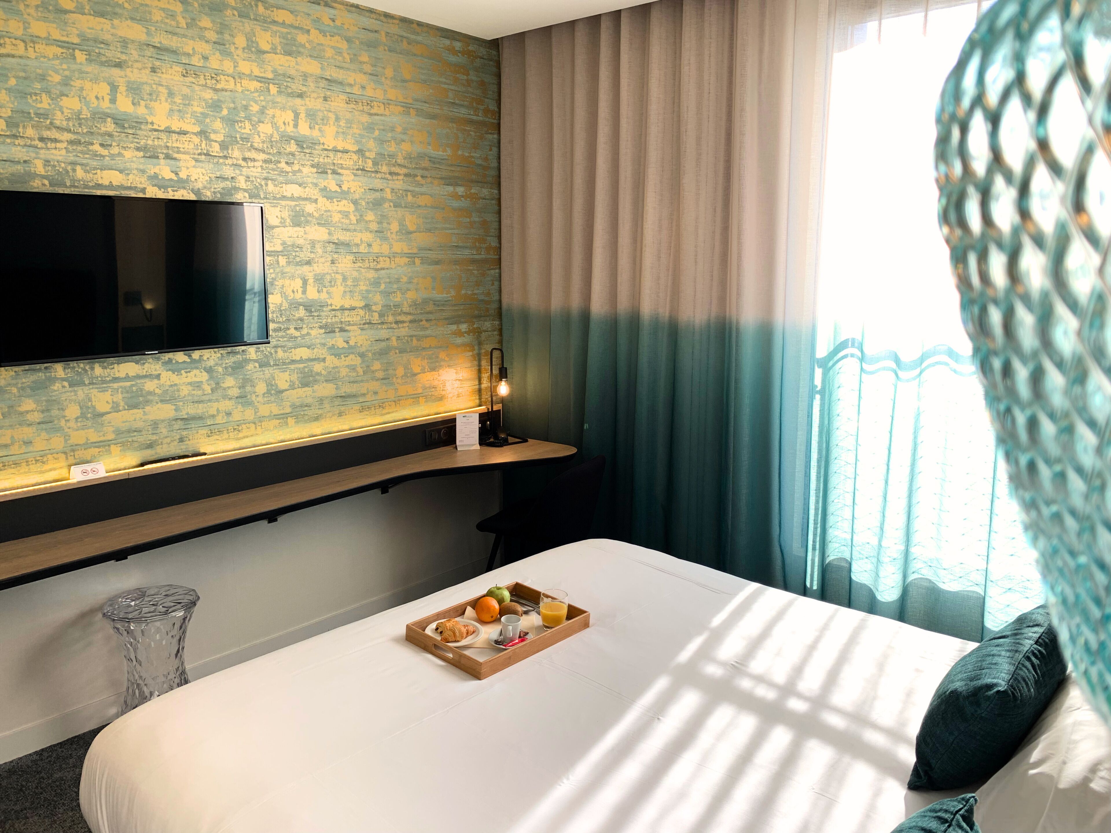 Quarto de hotel com cama, televisão de tela plana presa na parede, mesa de cabeceira com luminária e janela com cortinas.
