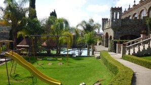 Um parquinho com escorrega amarelo e balanços, uma piscina e um prédio de pedra com arcos.