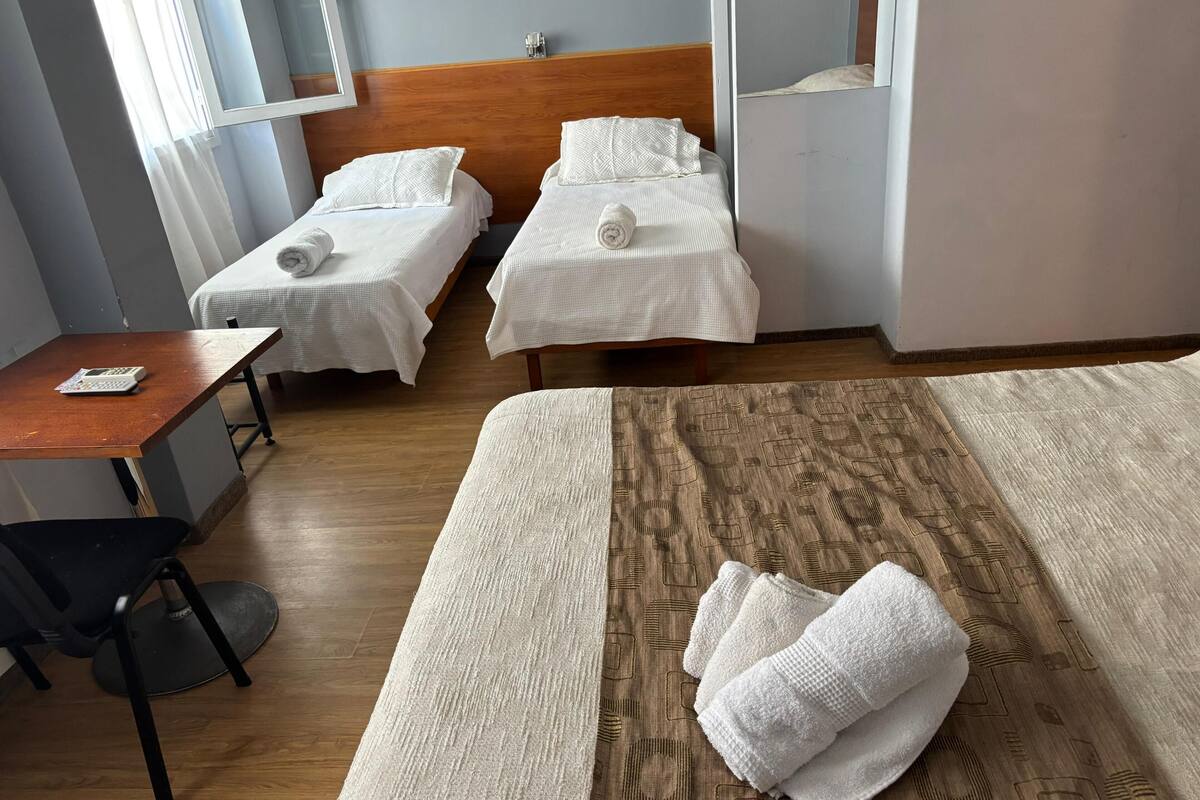 Quarto de hotel com duas camas de solteiro, uma escrivaninha de madeira, uma cadeira e uma cama grande com toalhas.