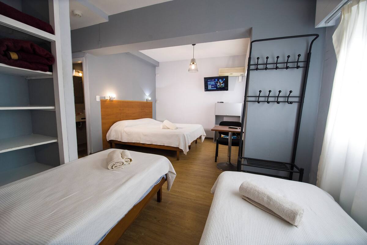 Quarto de hotel com duas camas de solteiro, cabeceira de madeira, escrivaninha com cadeira, TV fixada na parede e um cabideiro.