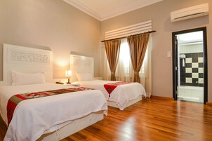 Quarto de hotel com duas camas, piso de madeira, porta do banheiro xadrez e um aparelho de ar condicionado.