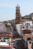 Vista da rua com uma torre do relógio imponente ao fundo, rodeada por edifícios com telhados de telhas vermelhas e carros estacionados ao longo da rua.