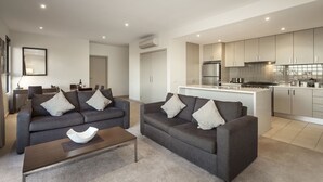 Uma sala de estar moderna com dois sofás cinzas, uma mesa de centro e uma área de cozinha com eletrodomésticos de aço inoxidável.