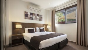 Quarto de hotel com uma cama grande, duas mesas de cabeceira com luminárias, um ar condicionado fixado na parede, uma janela com cortinas e um elemento decorativo sobre a cama.