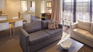 Quarto de hotel moderno com sofá, poltrona, área de refeições, cozinha compacta e grandes janelas com cortinas.