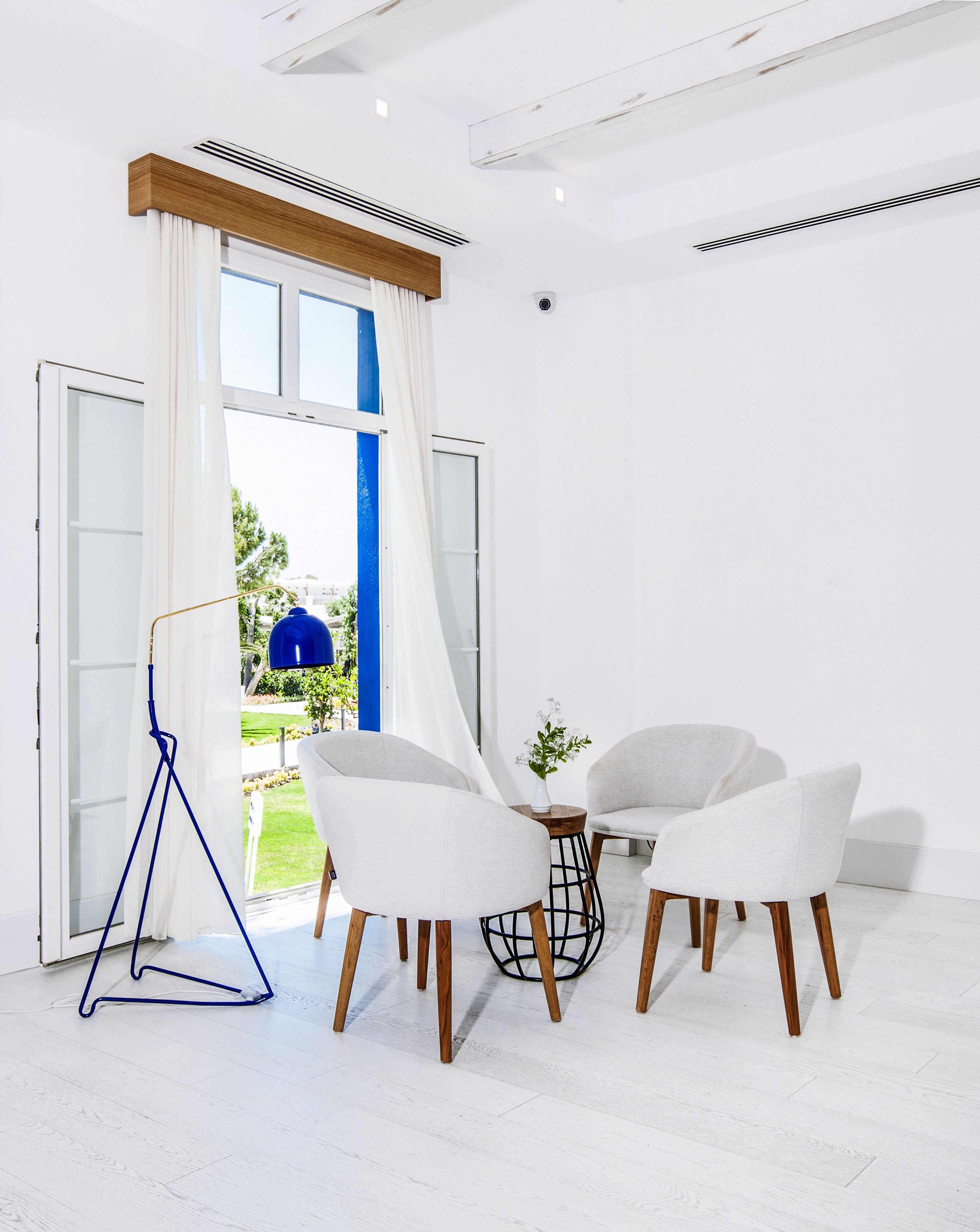 Quarto moderno com paredes brancas, uma janela grande com cortinas brancas, uma cadeira azul, uma mesa redonda e um abajur.
