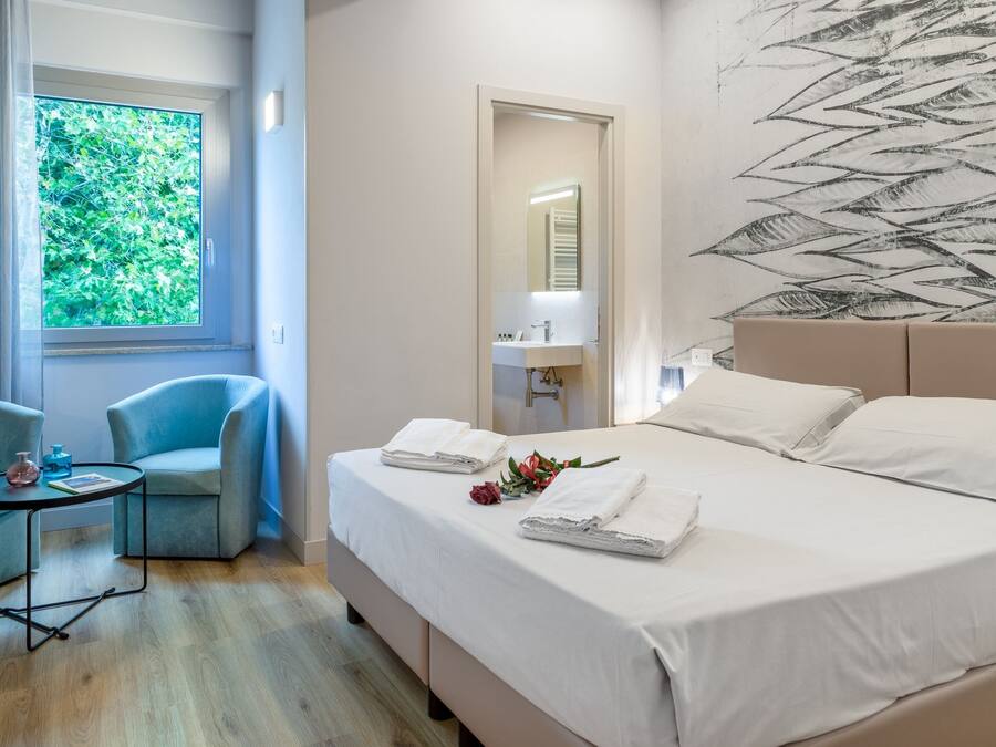 Quarto de hotel com cama, uma cadeira, uma mesa pequena, janela com vista para vegetação e parede com design abstrato em preto e branco.