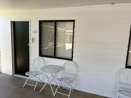 Fachada de prédio com porta fechada, janela com tela de metal e duas cadeiras de metal brancas.