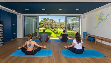 Um estúdio de yoga com quatro pessoas meditando em tapetes azuis, janelas amplas e vista para um parque.