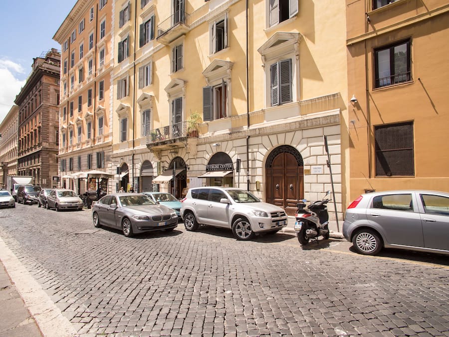 Uma rua de paralelepípedos flanqueada por edifícios históricos, carros estacionados e uma scooter.