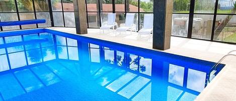 Piscina interior com água azul cristalina, rodeada por paredes de vidro e chão revestido de azulejos.