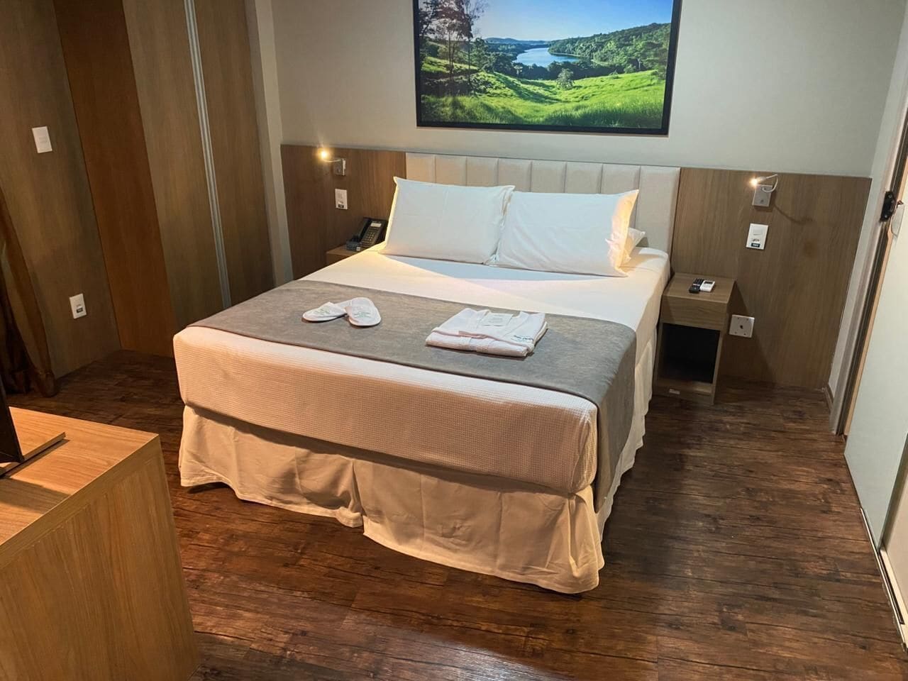 Uma cama bem arrumada com um edredom branco e travesseiros, uma mesa de cabeceira com abajur e uma janela grande com vista para uma paisagem verde.