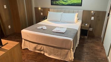 Uma cama bem arrumada com um edredom branco e travesseiros, uma mesa de cabeceira com abajur e uma janela grande com vista para uma paisagem verde.