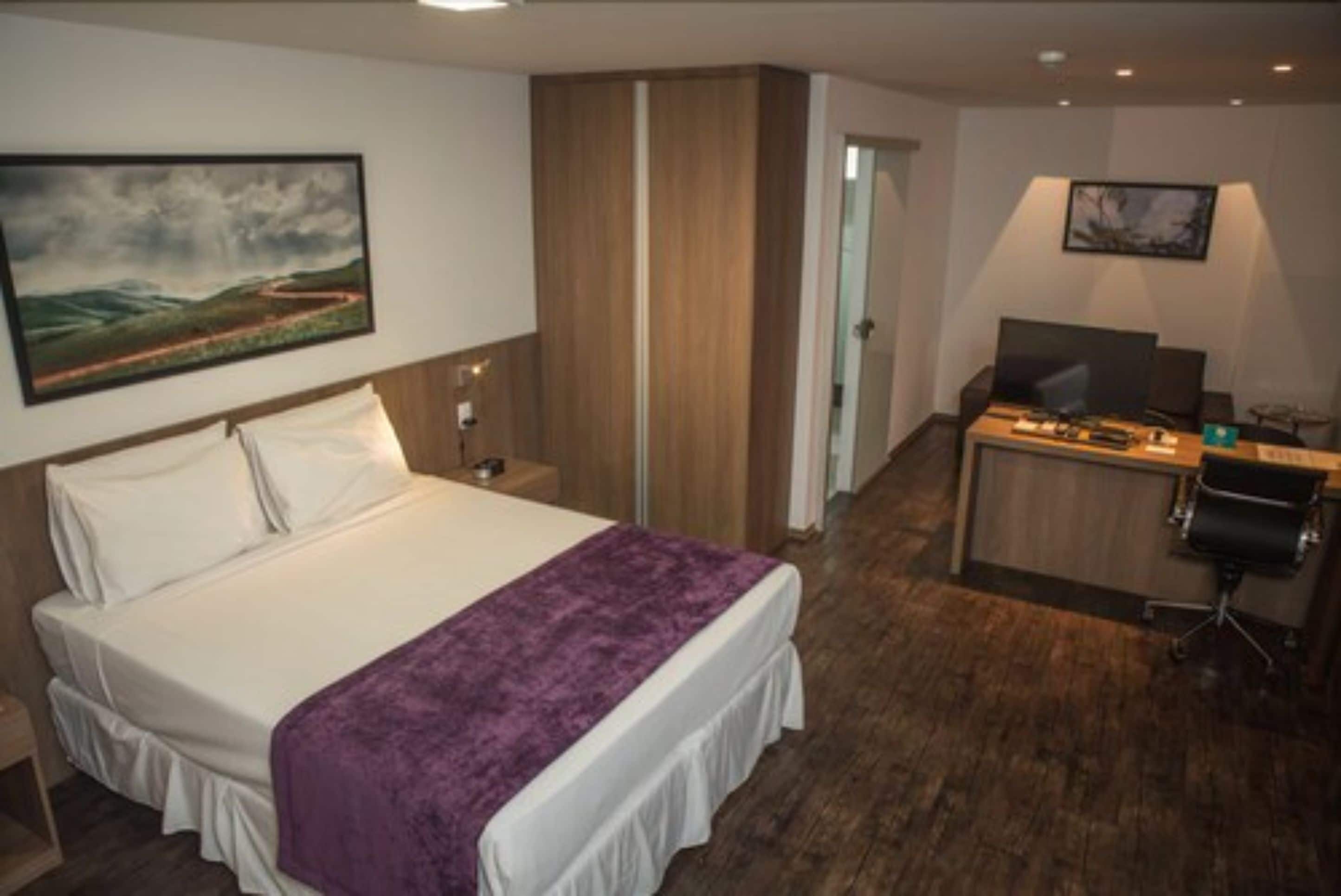 Quarto de hotel com cama, escrivaninha, cadeira e televisão.