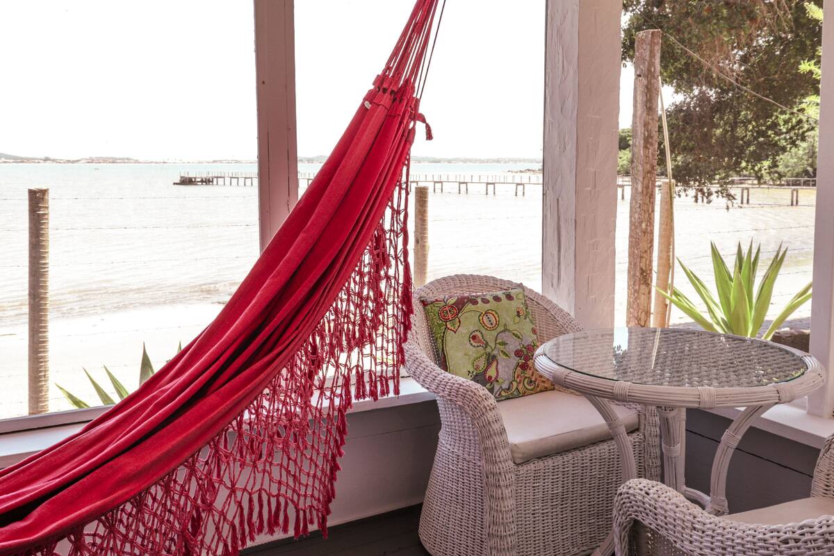 Conjunto de cadeira e mesa de vime em frente a uma janela com vista para uma praia e um balanço.