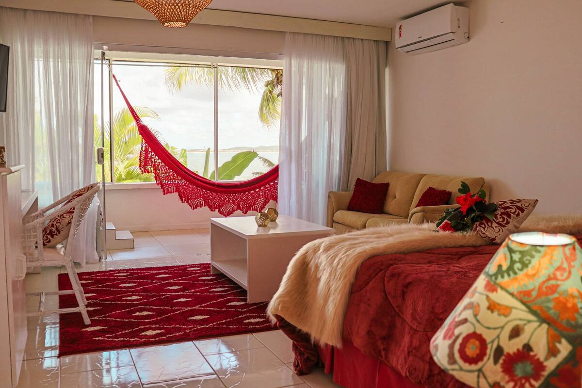Quarto com cama, rede vermelha, mesa de centro branca e vista para o exterior.