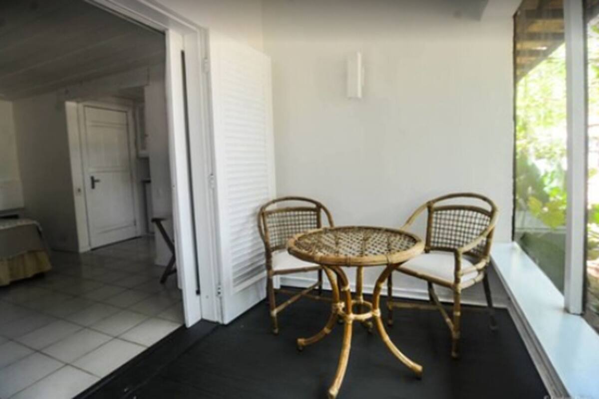 Varanda com mesa redonda e duas cadeiras de vime, porta branca e vista para o verde lá fora.